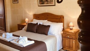 Quarto de hotel com cama de madeira, duas mesas de cabeceira com luminárias, um quadro na parede e vista do exterior por uma janela em arco.