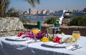 Mesa posta para dois com pratos, talheres e bebidas, com vista para um corpo d'água e horizonte urbano.