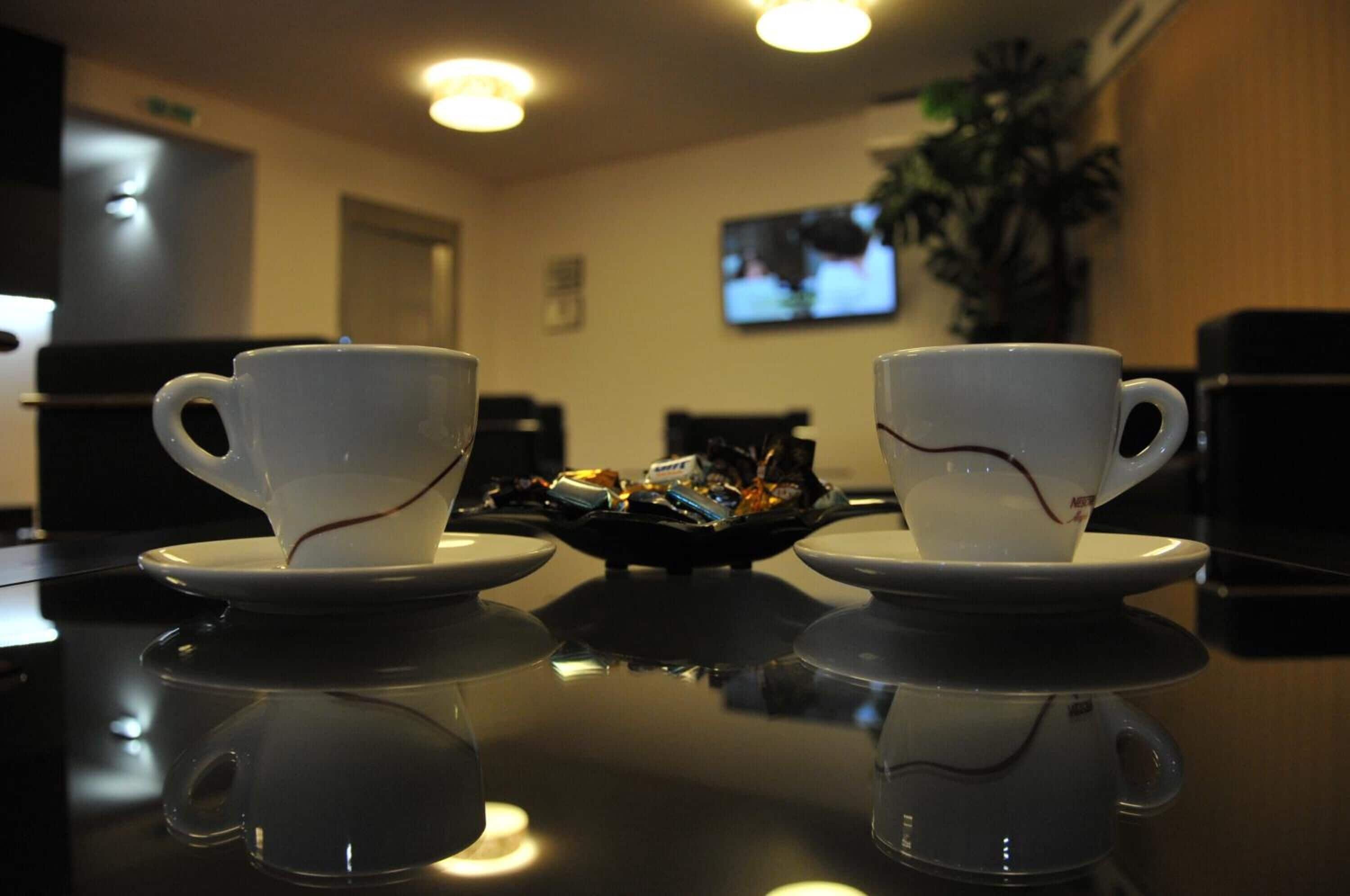 Duas chávenas brancas com desenhos castanhos sobre uma mesa espelhada, num quarto com uma televisão e uma planta ao fundo.