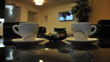 Duas chávenas brancas com desenhos castanhos sobre uma mesa espelhada, num quarto com uma televisão e uma planta ao fundo.