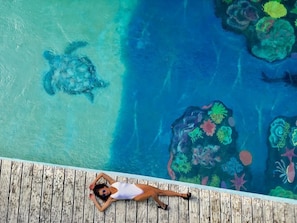 Mulher deitada em um deck de madeira perto de uma piscina com água cristalina e uma tartaruga nadando por perto.