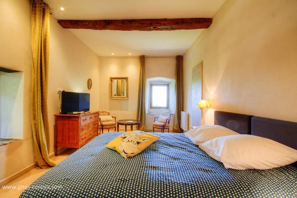Quarto de hotel com uma cama, uma escrivaninha com televisão, uma cadeira, uma mesinha e uma janela.