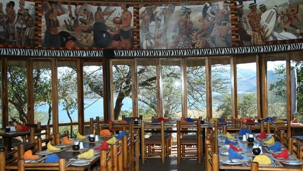 Um restaurante com mesas e cadeiras de madeira, janelas amplas e um mural na parede.