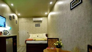 Quarto de hotel pequeno com uma cama, uma secretária com um computador, uma mesa com uma cadeira e um vaso de plantas.