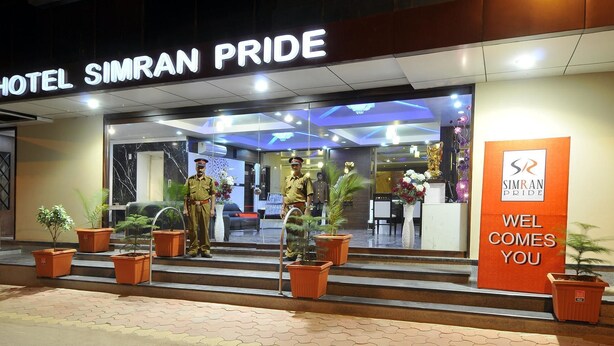 Exterior do Hotel Simran Pride, com entrada convidativa, vasos de plantas e um letreiro com a palavra 'Welcome'.