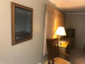Quarto de hotel com um espelho grande e detalhado, uma cadeira de madeira, uma escrivaninha com luminária e uma televisão.