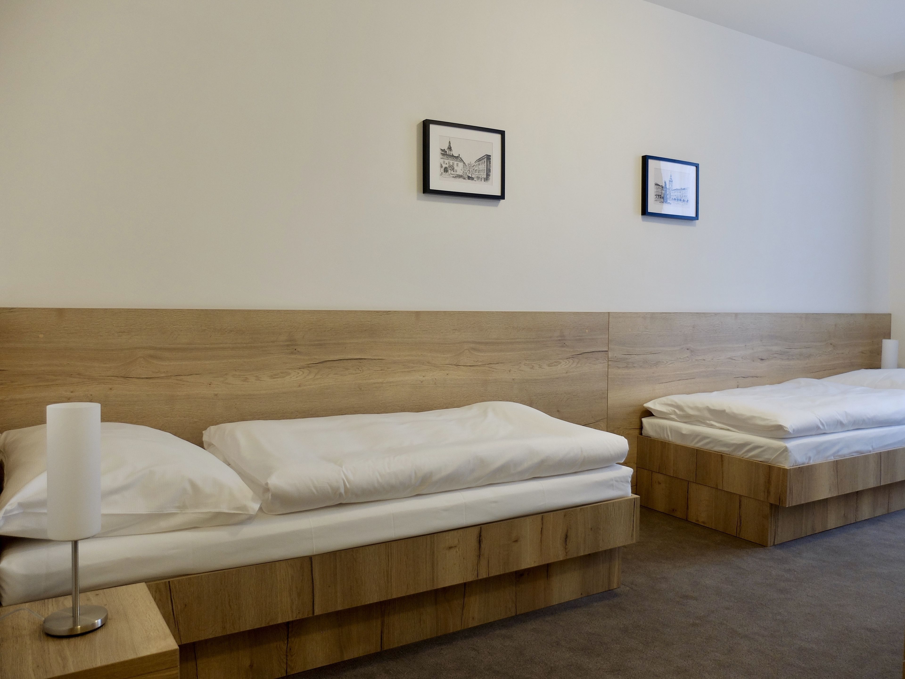 Quarto de hotel com duas camas de madeira, uma mesa de cabeceira com abajur e duas imagens emolduradas na parede.