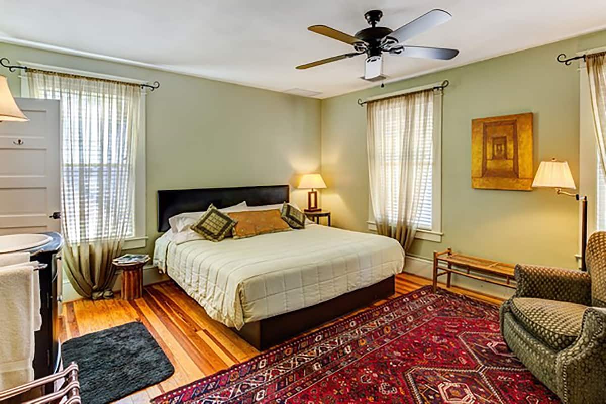Quarto com cama, ventoinha no teto, mesa de cabeceira, poltrona, tapete e um quadro na parede.