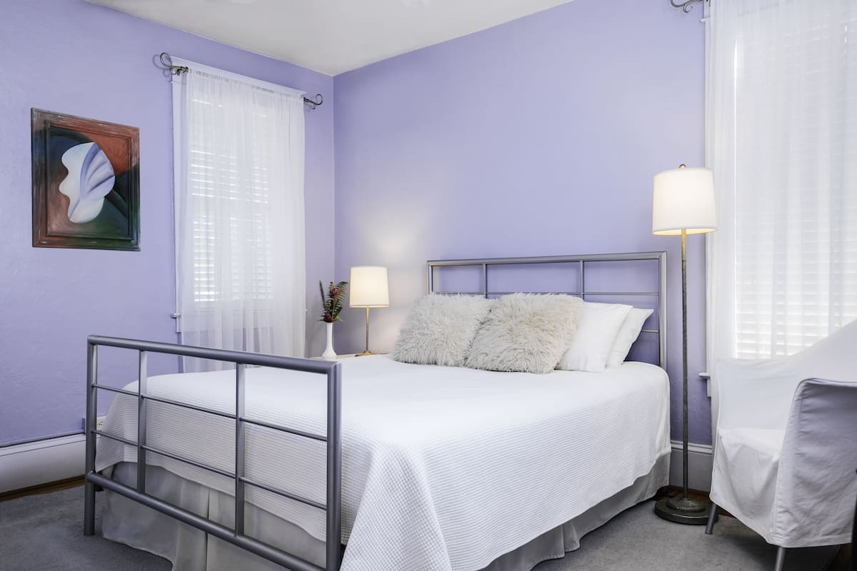 Quarto com estrutura de cama em metal, roupa de cama branca, mesa de cabeceira com abajur e uma cadeira.