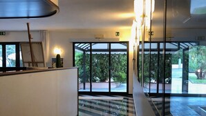 Um hall de hotel com uma mesa de recepção, uma escadaria que dá para área externa e grandes portas de vidro com vista para o exterior.