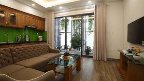 Uma sala de estar moderna com sofá seccional, uma mesa de centro de vidro e uma área de cozinha com armários de madeira e painel verde.