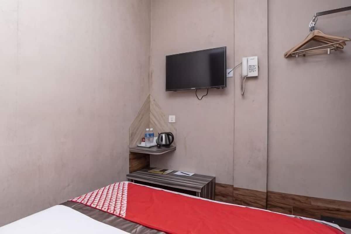 Quarto de hotel com uma cama, televisão presa na parede, uma mesinha com chaleira e casaco pendurado em um cabide.