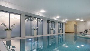 Uma piscina interior com água cristalina, rodeada por janelas com vidro jateado e arranjos de flores brancas.