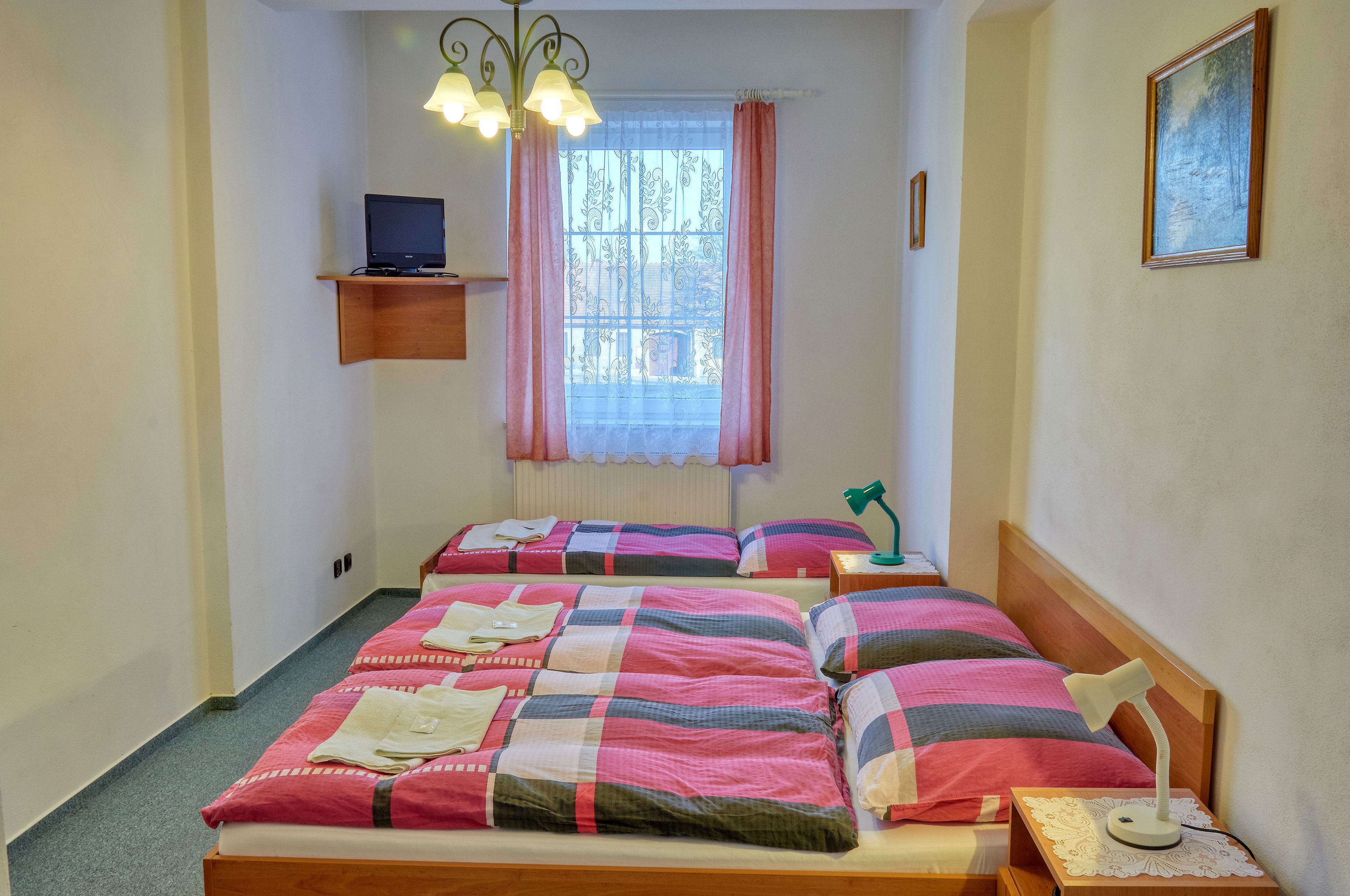 Quarto com duas camas, uma janela com cortinas, uma escrivaninha com luminária e uma pequena estante com televisão.