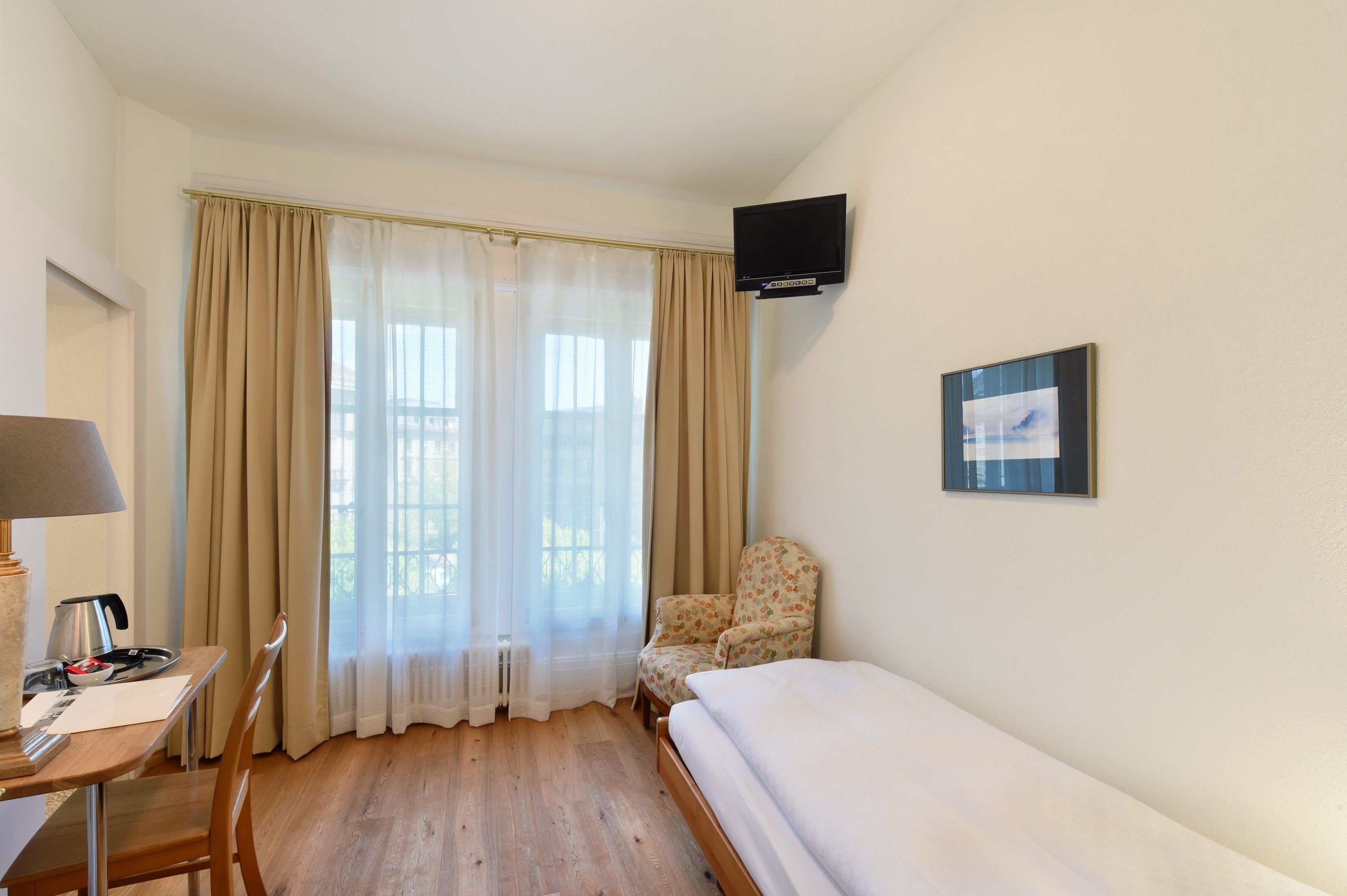 Quarto de hotel com cama, poltrona, escrivaninha com luminária, televisão e janela com cortinas.
