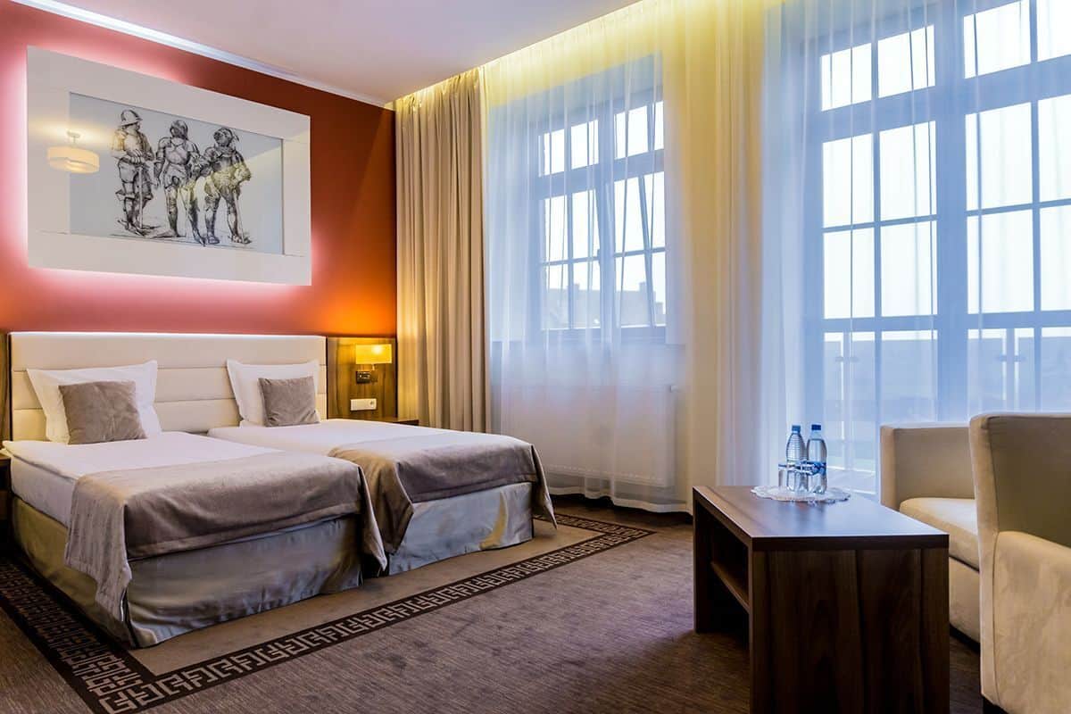 Quarto de hotel com duas camas, uma escrivaninha, uma cadeira, uma janela grande com cortinas e uma obra de arte na parede.
