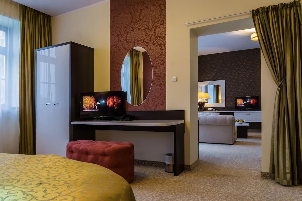 Quarto de hotel com cama, escrivaninha com computador, espelho e área de estar separada.