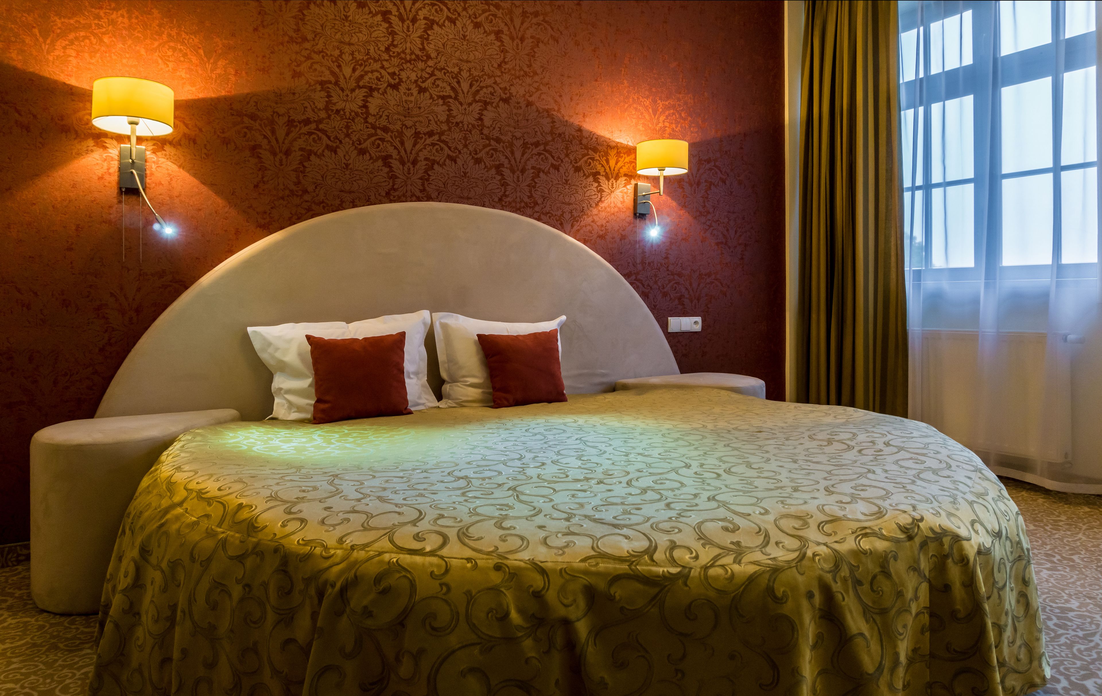 Quarto de hotel com uma cama grande, duas luminárias fixadas na parede, um painel de cabeceira curvado e uma janela com cortinas.