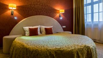 Quarto de hotel com uma cama grande, duas luminárias fixadas na parede, um painel de cabeceira curvado e uma janela com cortinas.