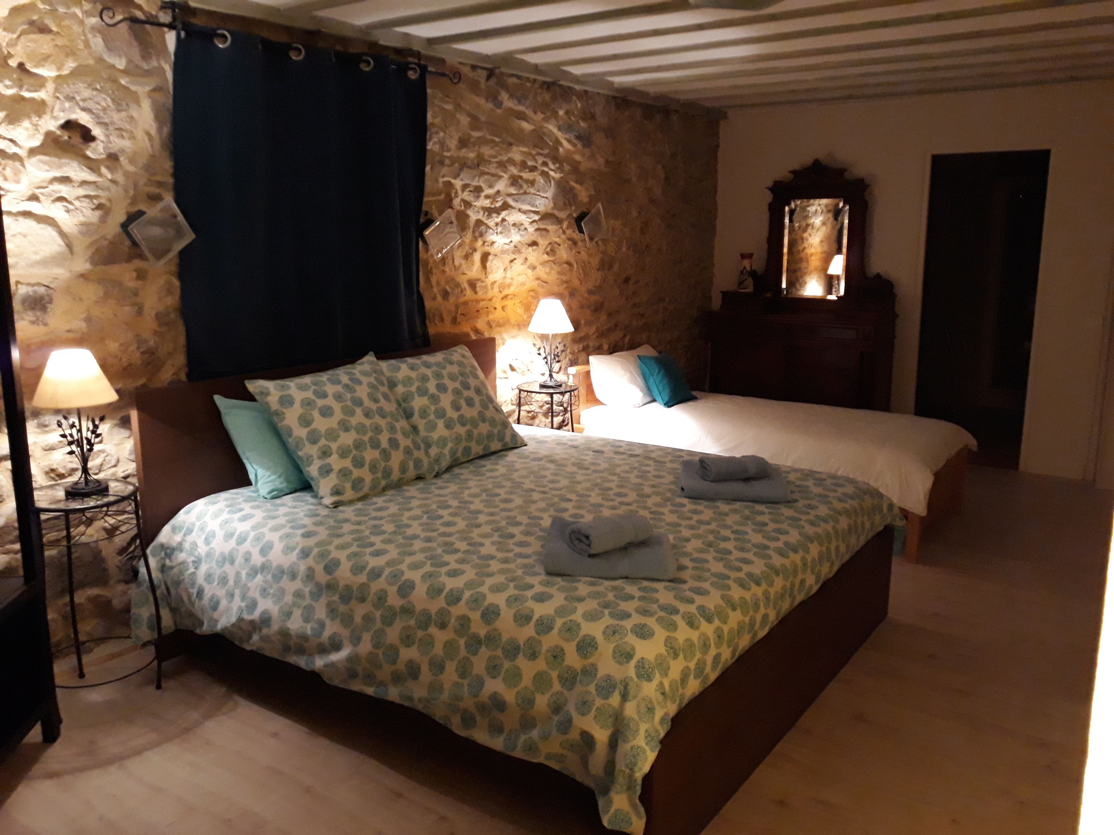 Quarto com parede de pedra, duas camas com roupa de cama estampada, mesa de cabeceira com abajur e cômoda.