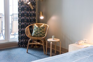 Quarto com cadeira de vime, um banquinho, mesa de cabeceira com abajur e janela com cortinas.