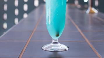 Um cocktail num copo alto com líquido azul, decorado com uma flor e uma fatia de fruta, sobre uma mesa com fundo estampado.