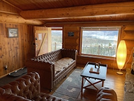 Interior acolhedor de uma cabana de madeira com um sofá de couro, uma mesa de madeira com peças de xadrez e vista para uma paisagem montanhosa coberta de neve através de grandes janelas.