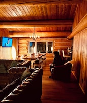 Interior acolhedor de uma cabana de madeira, com um sofá de couro, mesa de jantar, cadeiras e uma televisão.