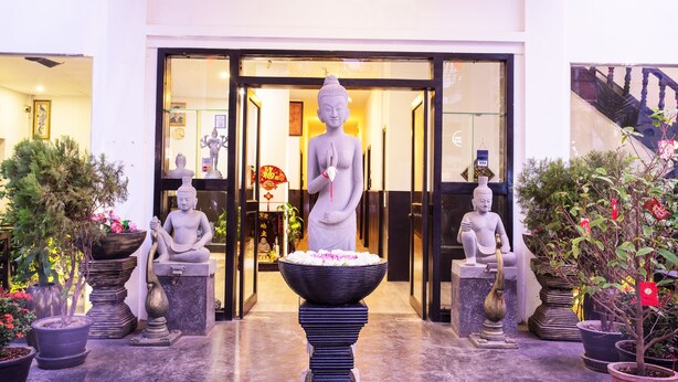 Estátua de uma figura sentada dentro de uma grande taça, acompanhada por outras duas estátuas, com plantas em vasos e a entrada de um edifício ao fundo.