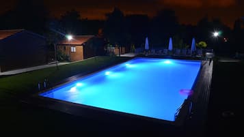 Uma piscina retangular iluminada com luz azul à noite, rodeada por um deck de madeira e um pequeno abrigo.