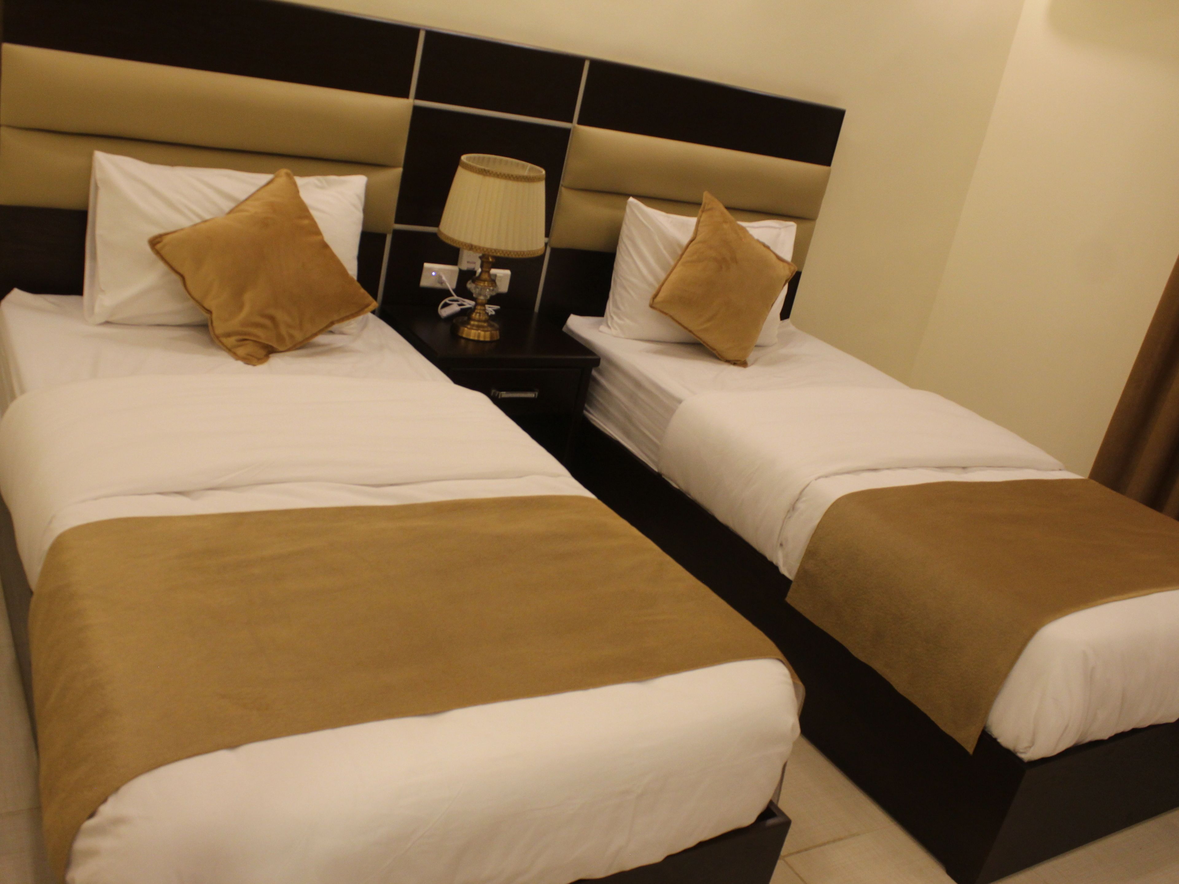 Quarto de hotel com duas camas, cada uma com roupa de cama branca e mantas marrons. Há uma mesa de cabeceira com abajur e telefone entre as camas.
