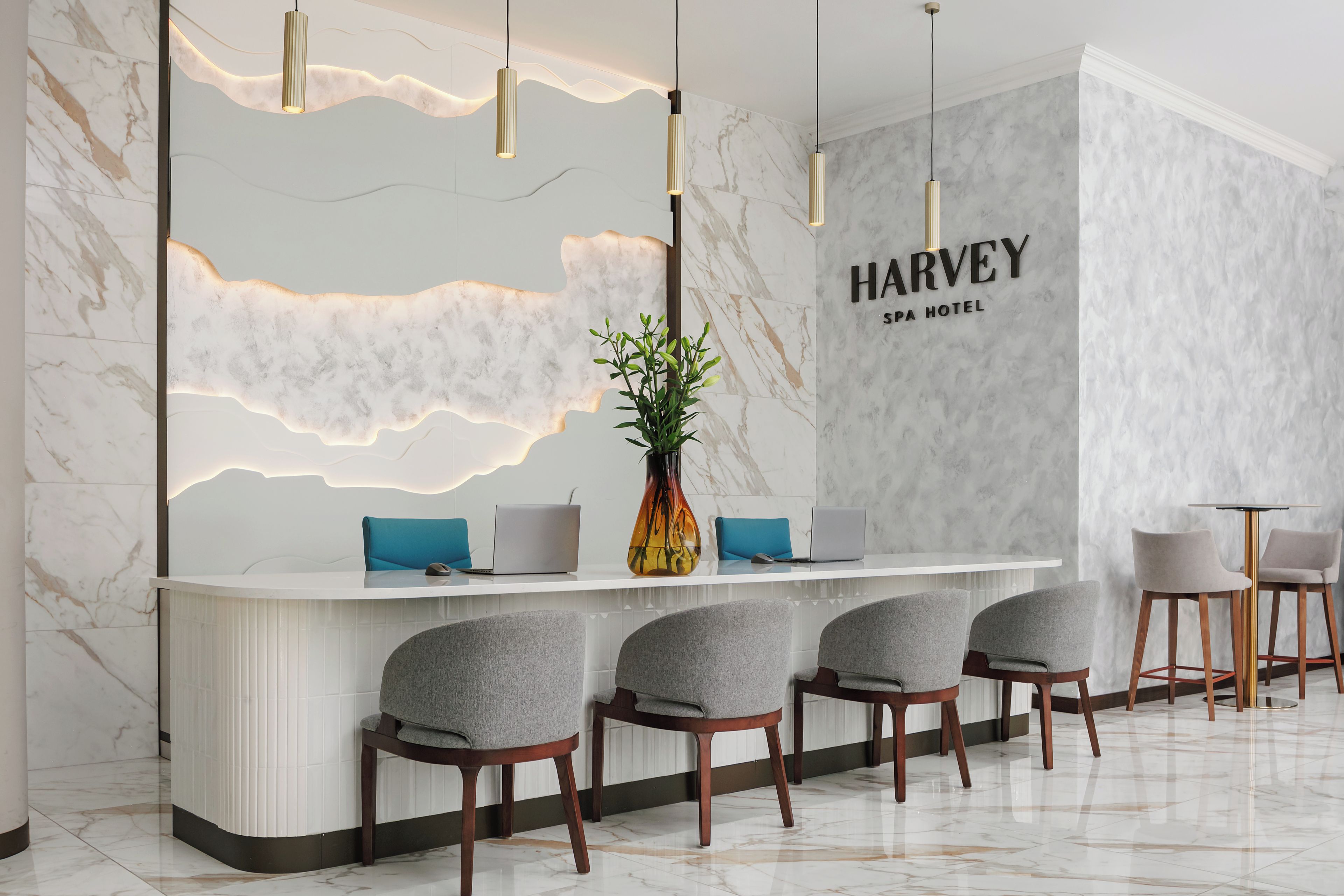 Uma área de recepção com parede de mármore, placa do Harvey SPA HOTEL, balcão de recepção e quatro cadeiras cinzentas.