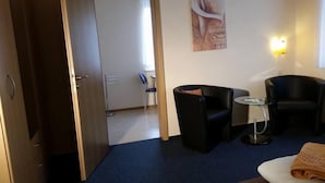 Quarto de hotel com porta de madeira escura, tapete azul, duas poltronas pretas, uma mesa redonda pequena e um quadro na parede.