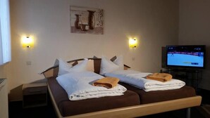 Uma cama de casal com roupa de cama branca e castanha, duas mesas de cabeceira, uma TV fixada na parede e duas arandelas.