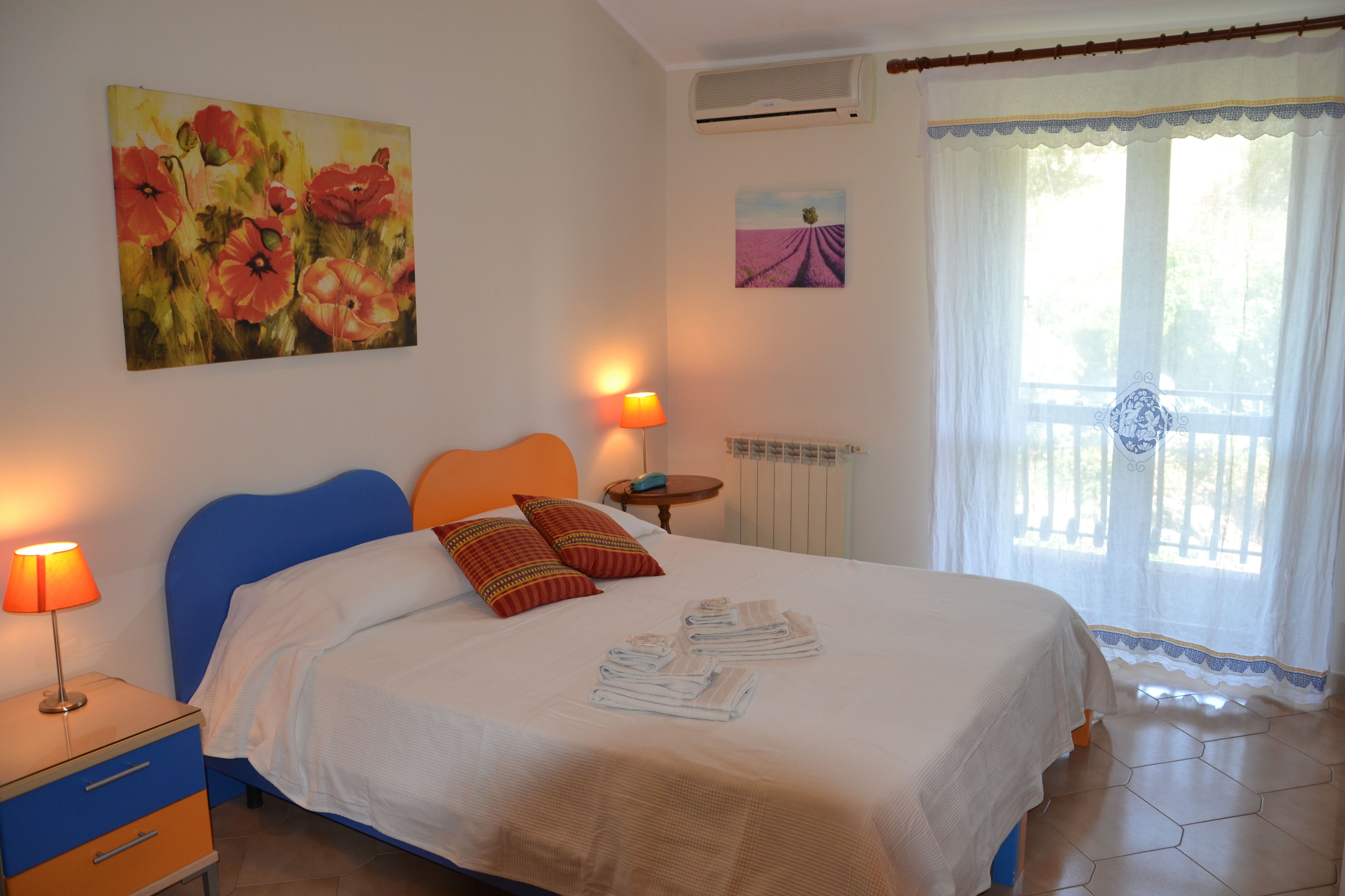 Um quarto com cama, abajures, um quadro, radiador e janela com cortinas.