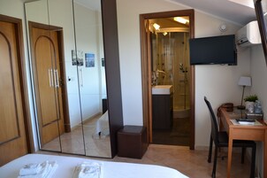 Quarto de hotel com cama, escrivaninha, cadeira, televisão, espelho e banheiro visível através de uma porta aberta.