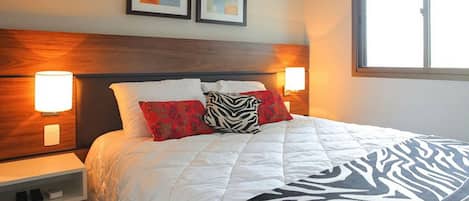 Quarto de hotel com cama grande, duas luminárias de cabeceira, uma cabeceira e duas imagens emolduradas na parede.