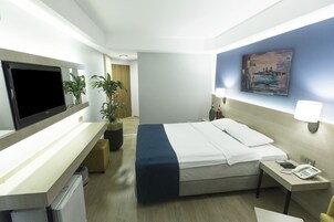 Quarto de hotel com cama, mesa de cabeceira com abajur, televisão e um quadro na parede.