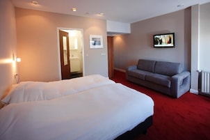 Quarto de hotel com uma cama, um sofá cinzento, uma televisão e um tapete vermelho.