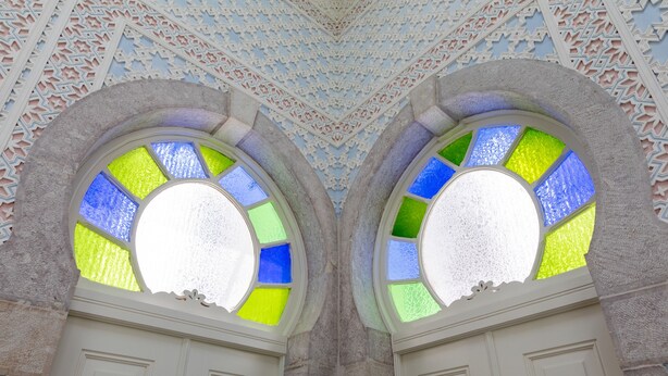 Duas janelas em arco com vitrais coloridos, embutidas numa parede de pedra com esculturas detalhadas.