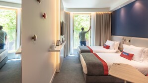 Quarto de hotel com duas camas, uma mesa, uma televisão e janela com cortinas.