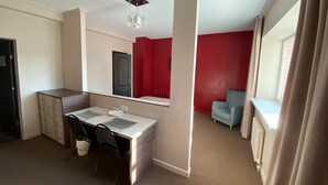 Quarto compacto com cozinha compacta, uma mesa com duas cadeiras, uma parede de destaque vermelha, uma janela com cortinas e um radiador.