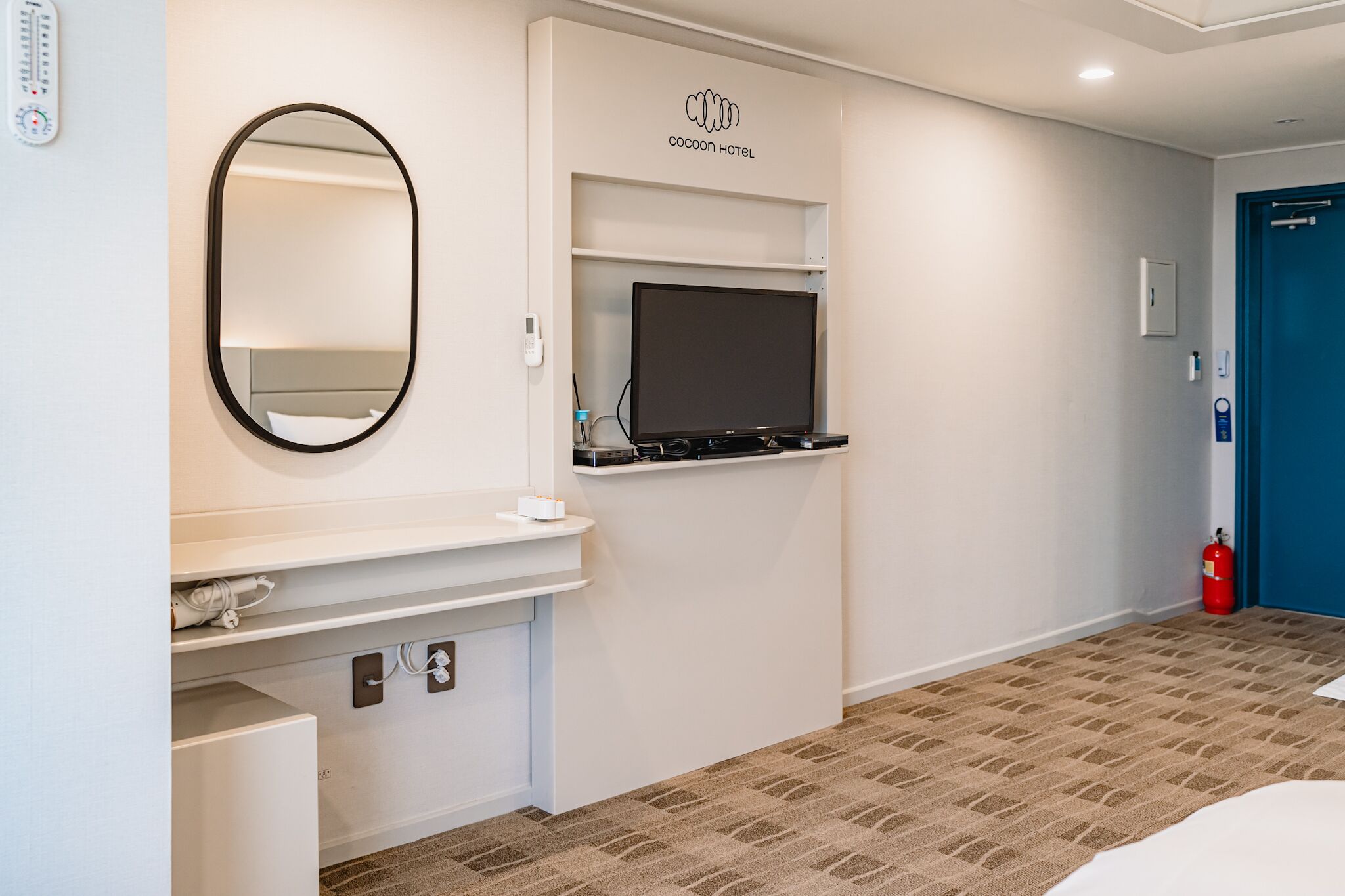 Quarto de hotel com uma toucador branco, uma televisão montada, uma placa Cocoon Hotel, uma porta azul e um extintor de incêndio.