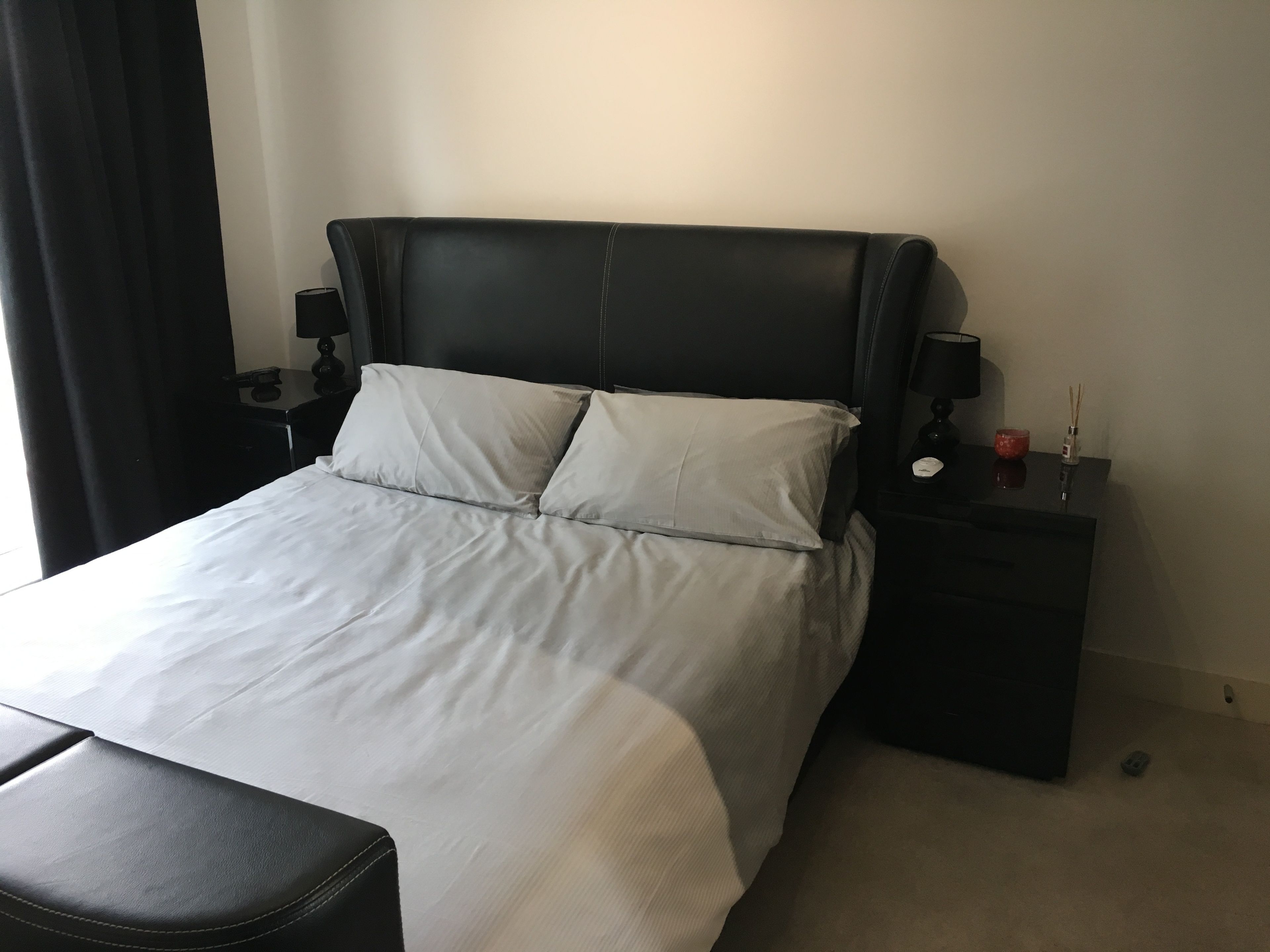 Cama bem arrumada com cabeceira preta, duas mesas de cabeceira com abajures e uma mesinha com um objeto vermelho.