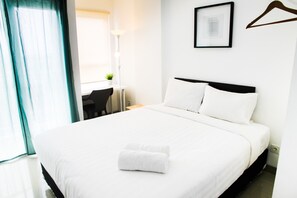 Uma cama bem arrumada com roupa branca e toalha enrolada, uma escrivaninha com cadeira e um cabideiro.