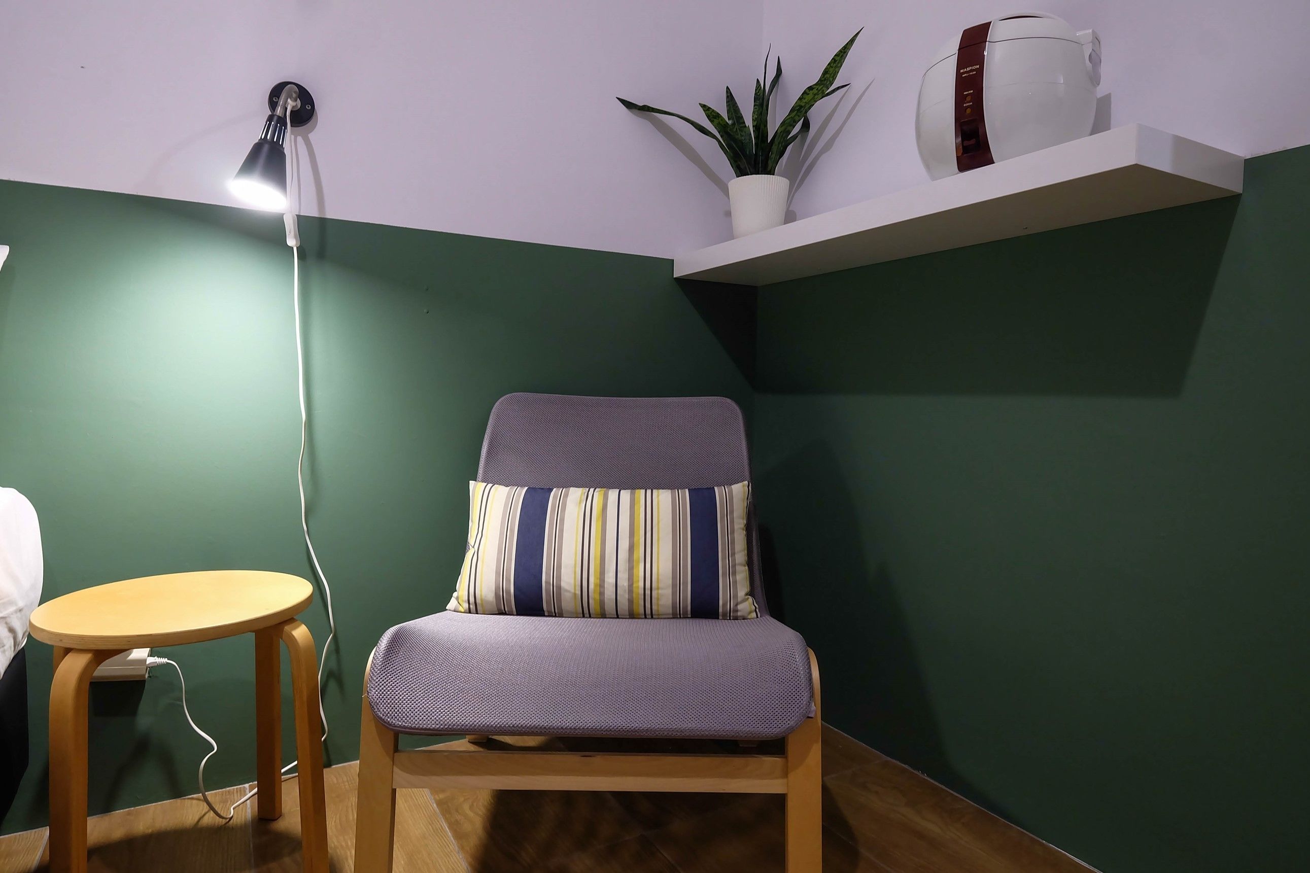 Quarto nos fundos com parede verde, uma cadeira de madeira com almofadas listradas, uma pequena mesa redonda, abajur e prateleira branca com uma planta e um objeto branco.