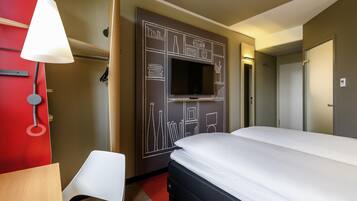 Quarto de hotel com cama, televisão na parede, secretária com candeeiro e uma cadeira.