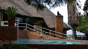 Uma área de piscina com um edifício de telhado de palha, uma chaminé de tijolo e uma escadaria que leva a uma varanda.