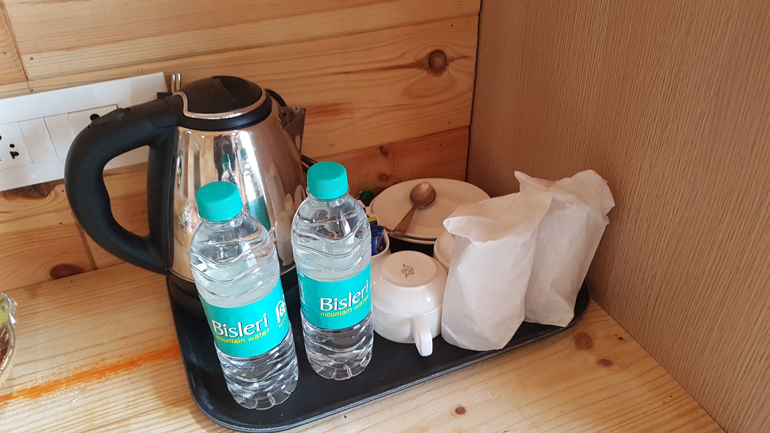 Uma cozinha compacta com chaleira, garrafas de água mineral Bisleri, uma caneca branca e toalhas de papel.