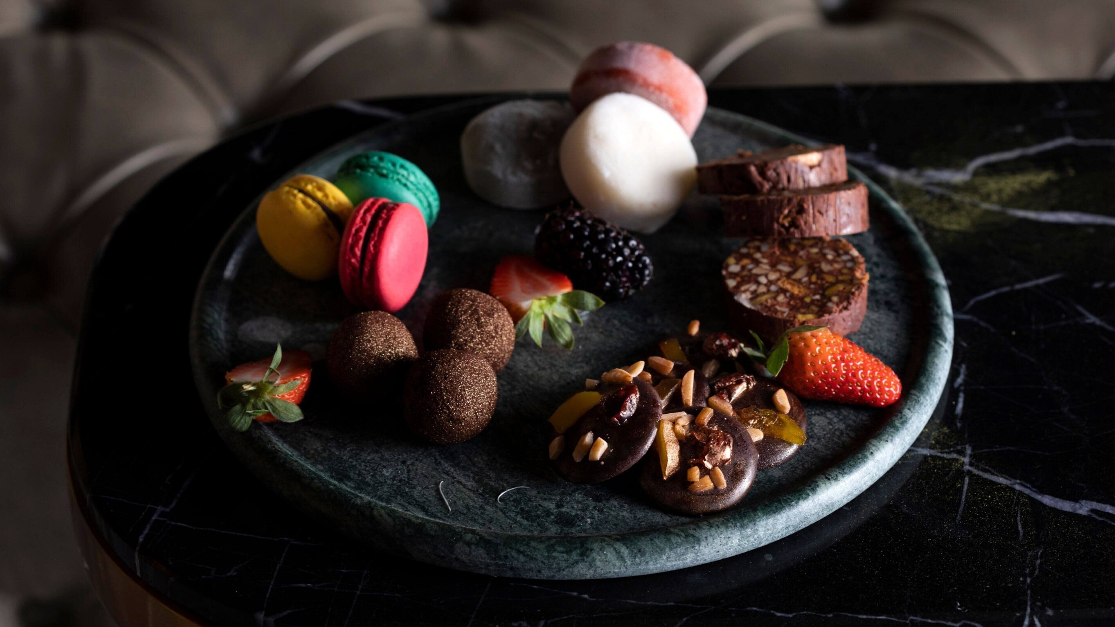 Uma travessa de sobremesas com macarons, trufas de chocolate, morangos e um doce com nozes cobertas de chocolate sobre uma mesa de mármore escuro.